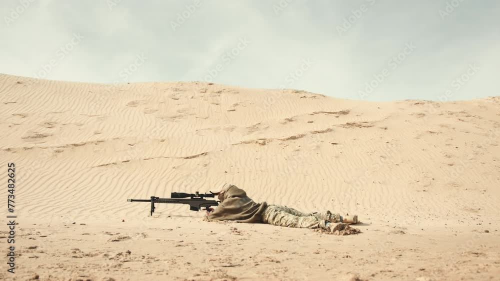 A sniper in desert camouflage lies prone with a rifle on a sand dune ...