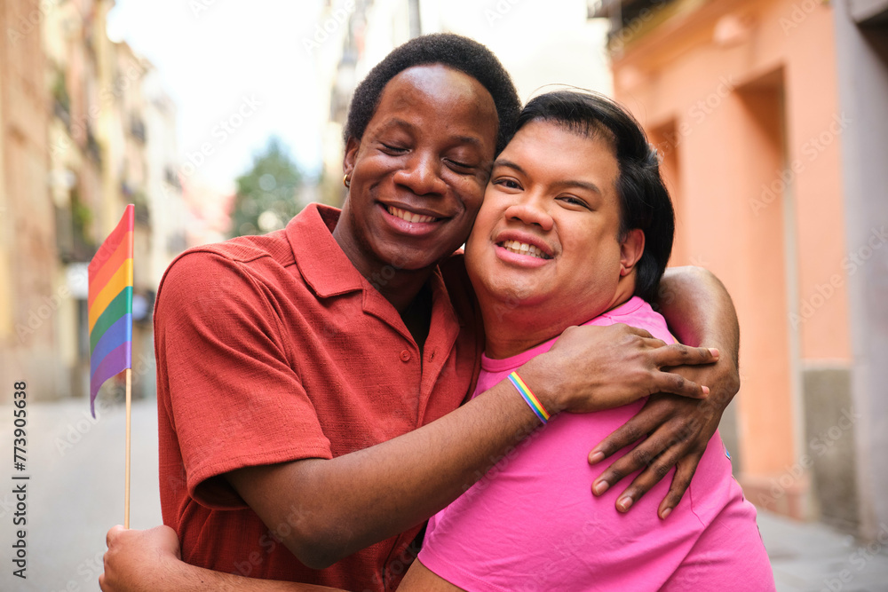 Gay multiracial couple hug each other while one of them holds a rainbow ...