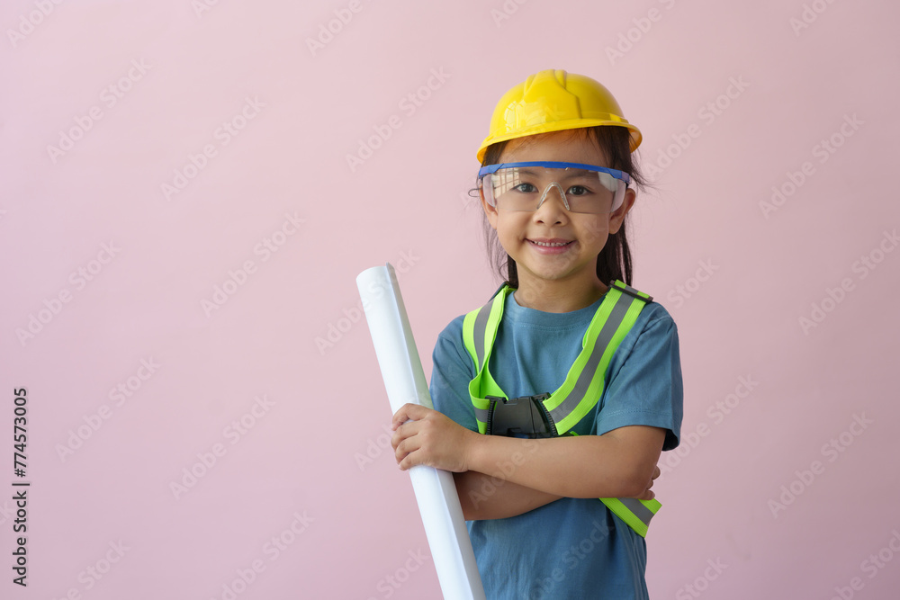 A cute girl wearing a yellow construction hat or safety helmet standing on a pink background ...