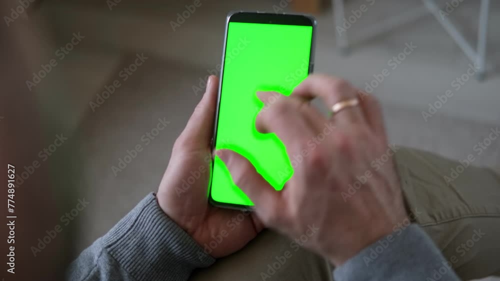 Young man sitting at home holding smartphone green mock-up screen in ...