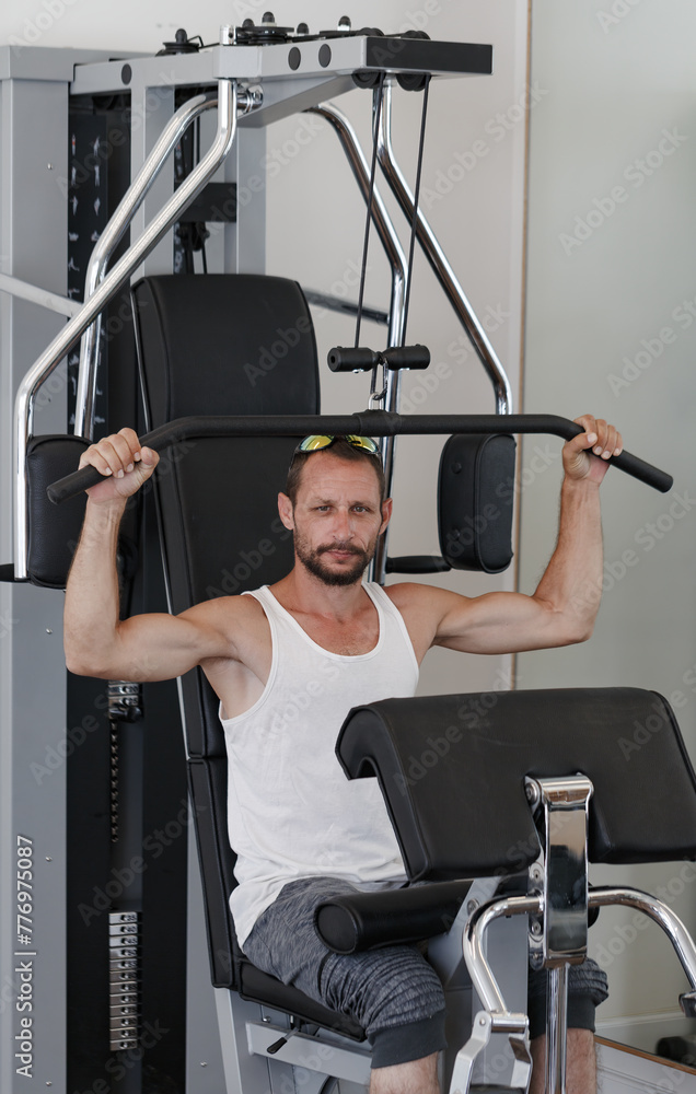 European man using a gym machine pulling down a bar attached to weights ...