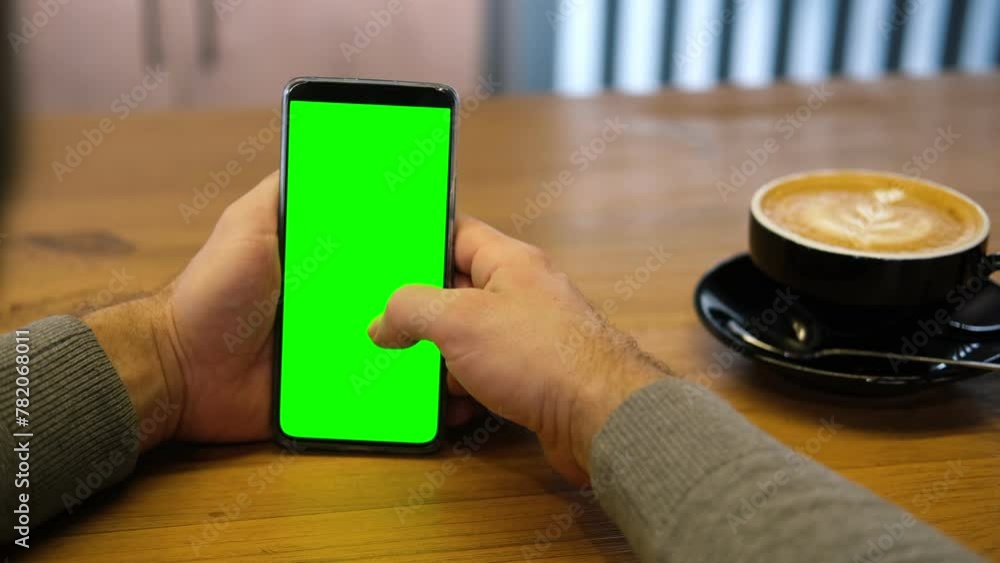 Young man sitting at cafe holding smartphone green mock-up screen in hand. Male person using ...