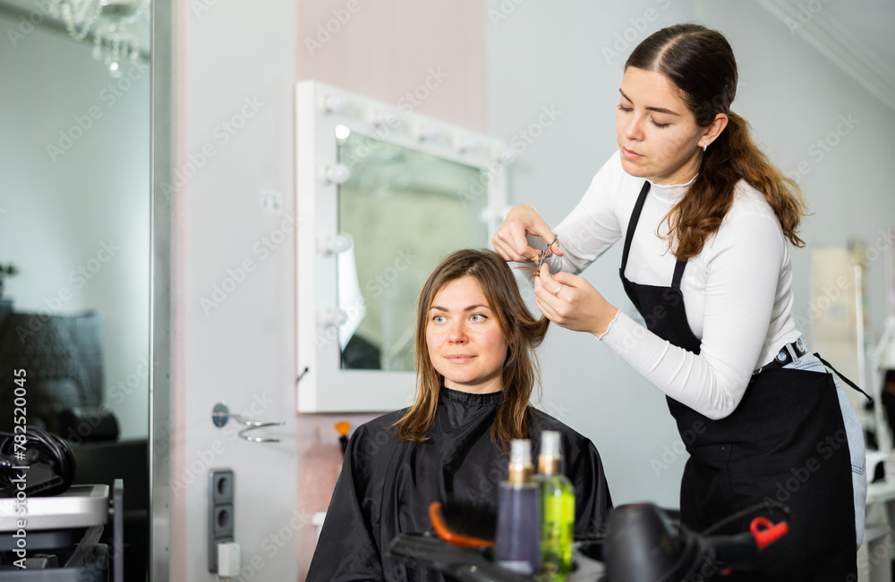 Focused female hairdresser cuts the hair of a young woman client with ...