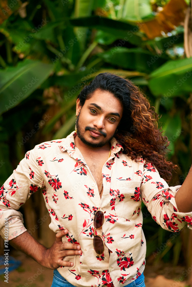 Confident gay man with wavy hair poses in tropical garden, vibrant ...