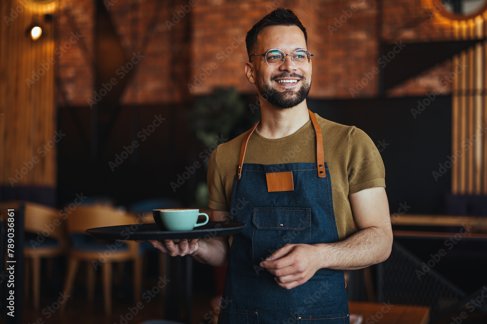 Happy waiter holding a tray while working in cafe and looking at camera ...