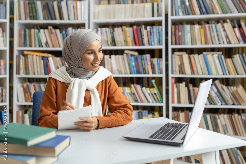 Smiling Muslim woman in hijab studying with a laptop and writing in a ...