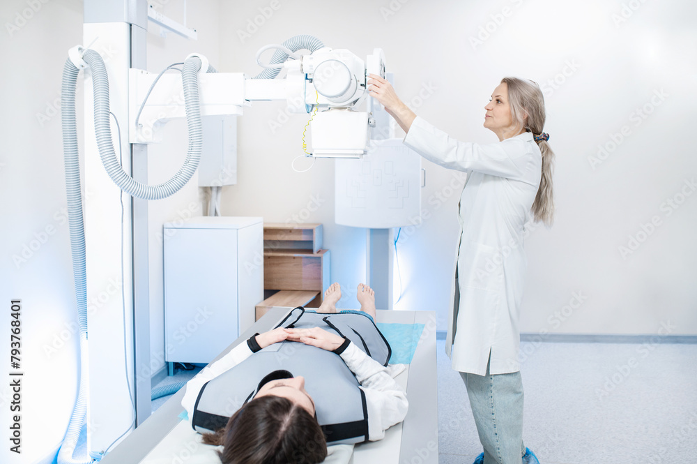 Female patient lying on bed while female nurse adjusting modern X-ray machine for scanning her ...