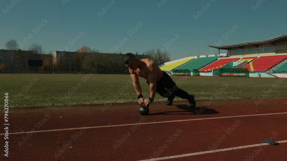 Young man is training at city stadium and doing push-ups on kettlebell ...