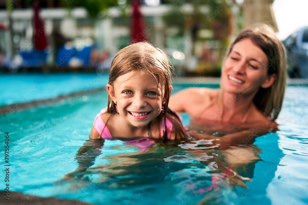 Smiling girl in pink swimsuit learns swimming in pool, mother supports ...