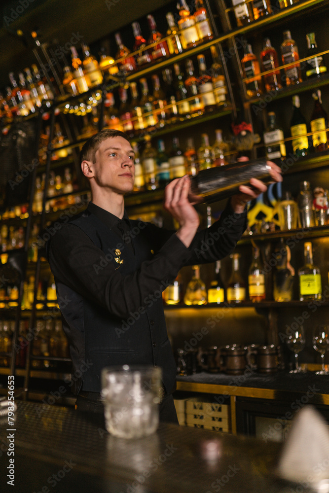 Professional barman in uniform shakes cocktail by counter. Serious ...
