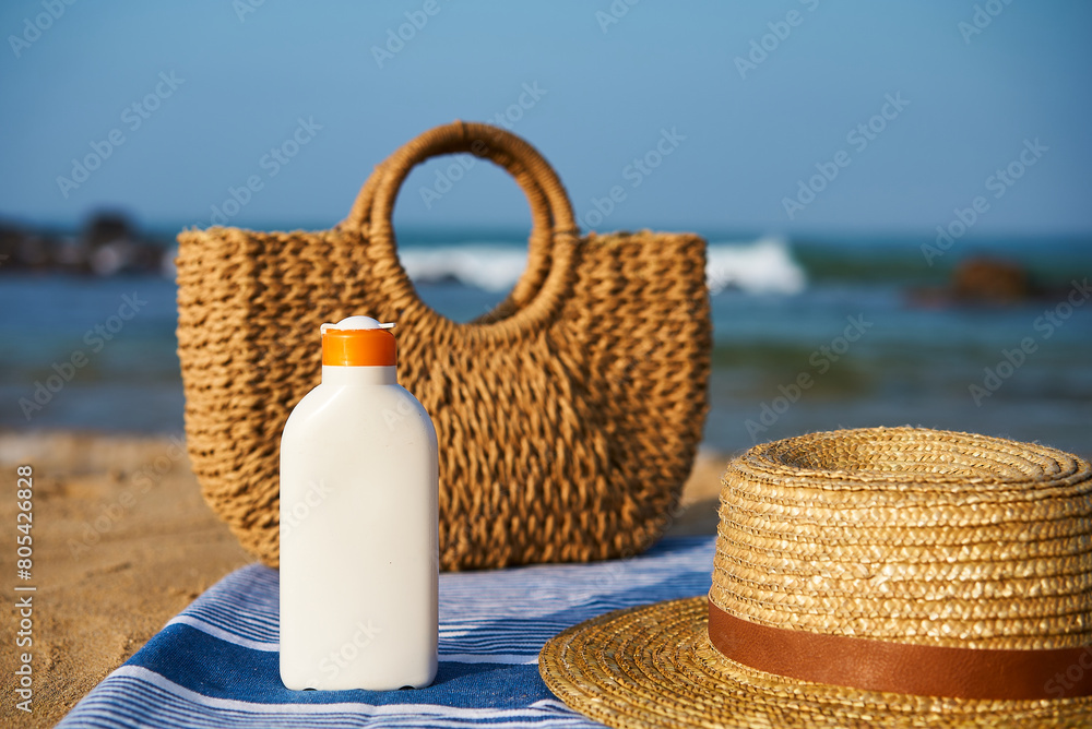 Wicker bag, sun hat, sunscreen bottle on striped towel at sunny seaside ...
