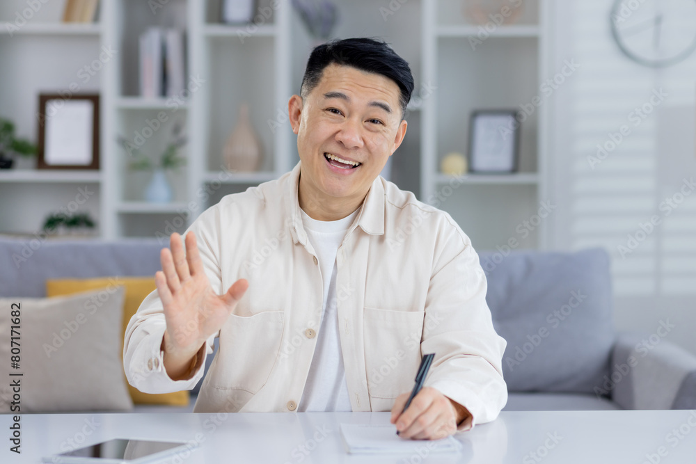 Cheerful Asian man sitting at home, talking and waving to the camera ...