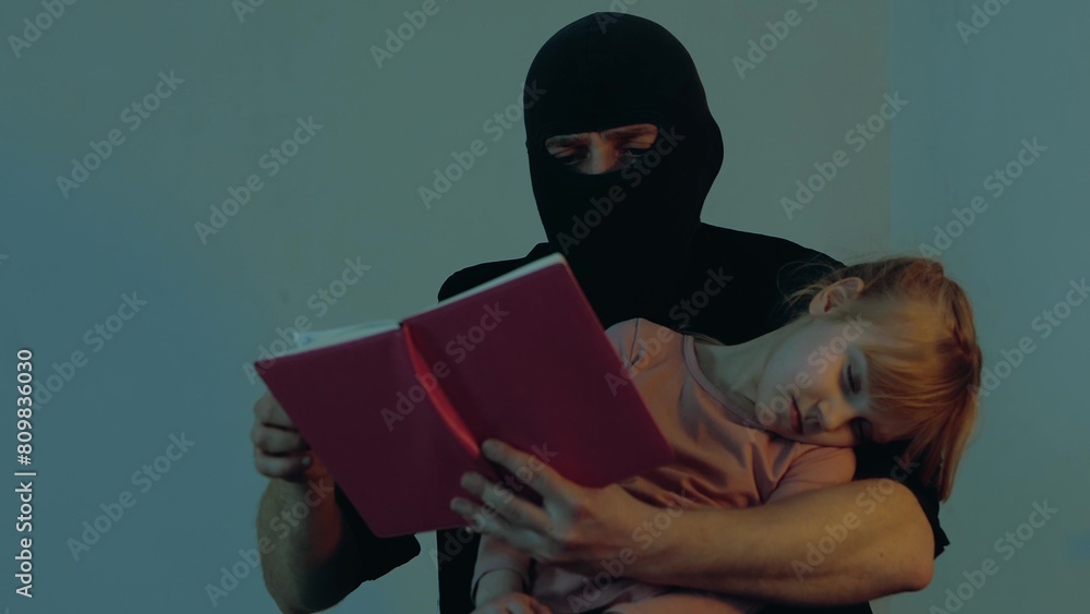 Kidnapper in a black balaclava holding a child in his arms, using book ...