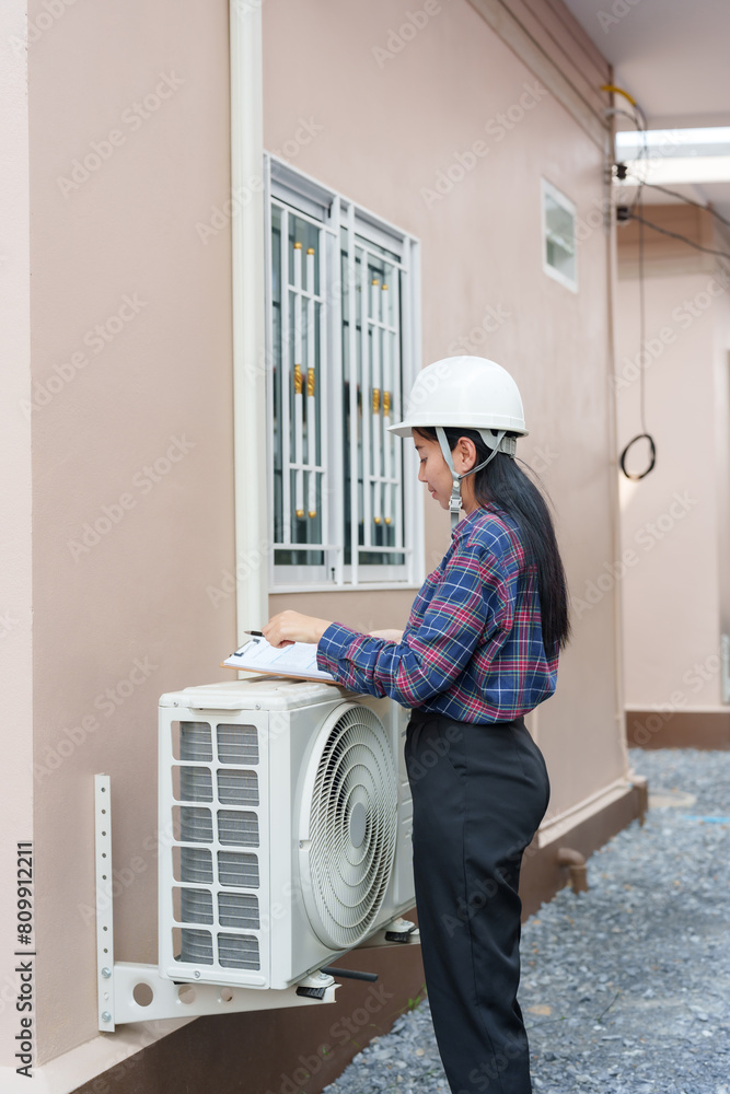 Asian professional in safety helmet writing on clipboard by air con ...