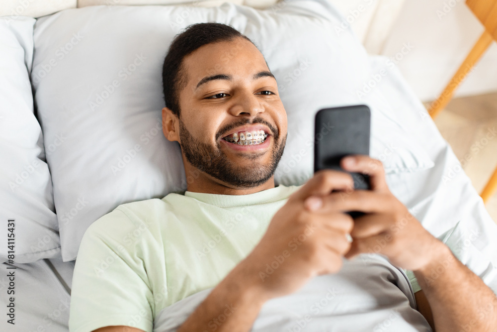 African American man is lying down in a bed, holding a cell phone in his hand. He appears to be ...