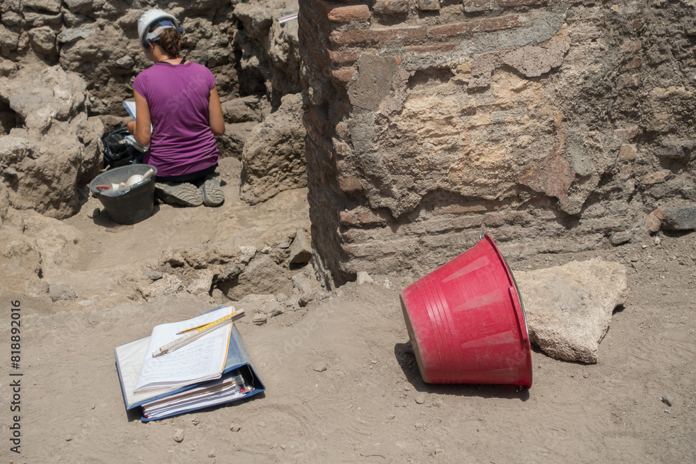 A female archaeologist kneels in a ancient site wearing a hard hat.In ...