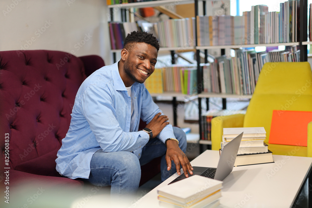 Young man studying in college library, sitting with laptop, surrounded ...