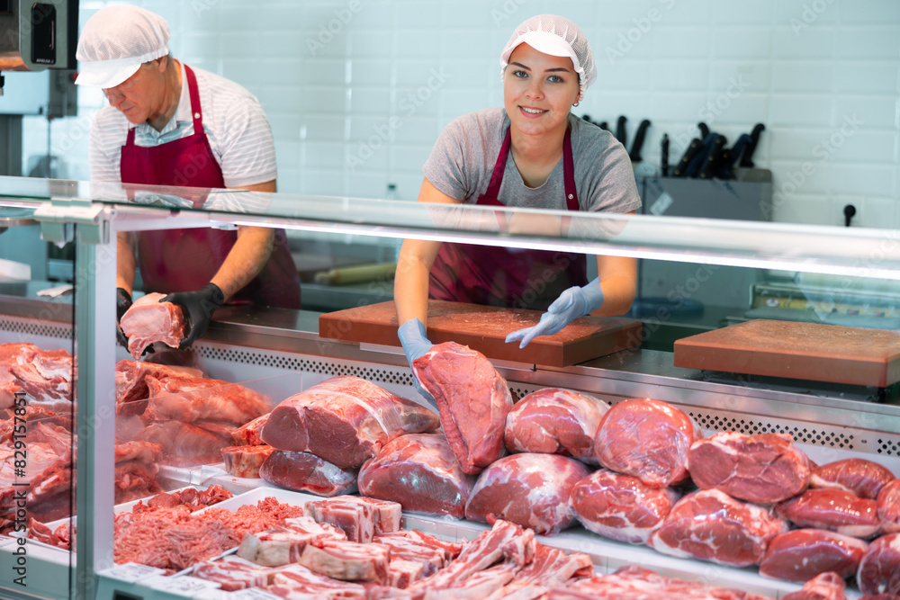 Smiling skilled young female butcher working behind counter in butchery ...
