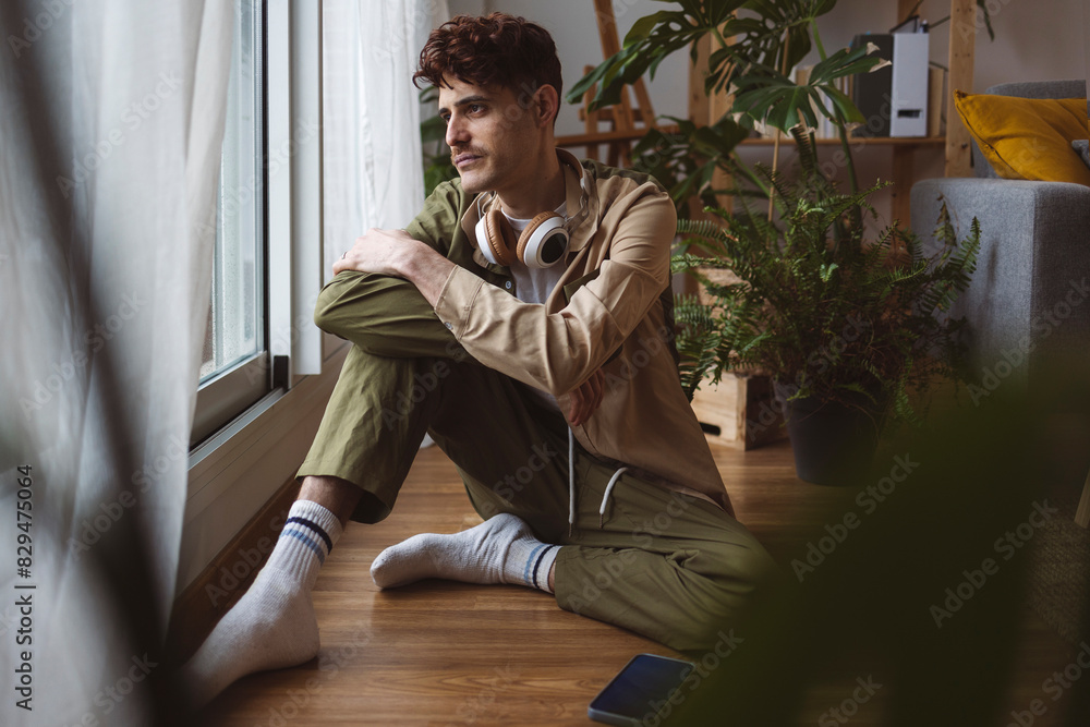 Thoughtful man sitting on floor and looking through window at home ...