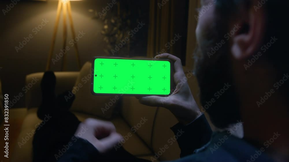 Man lying on couch using smartphone horizontally with chroma key green screen, scrolling through ...