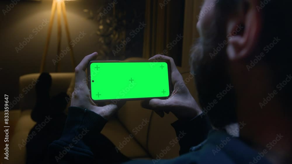 Man lying on couch using smartphone horizontally with chroma key green screen. Male watching ...