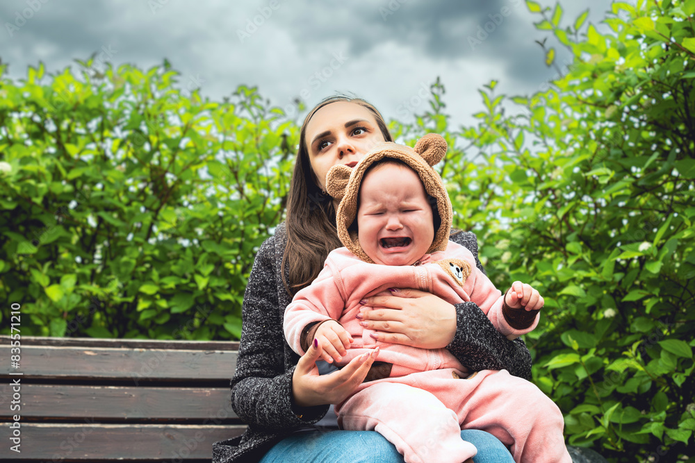 A tired mother sits on a park bench with her baby who is crying ...