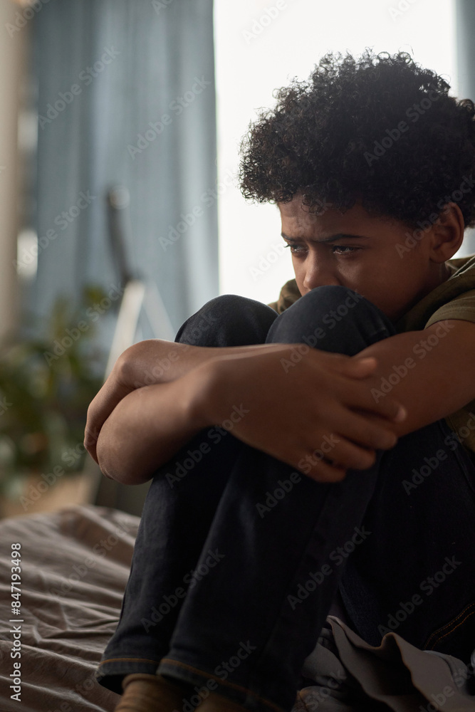 Vertical portrait of crying young boy sitting on bed in his room and ...
