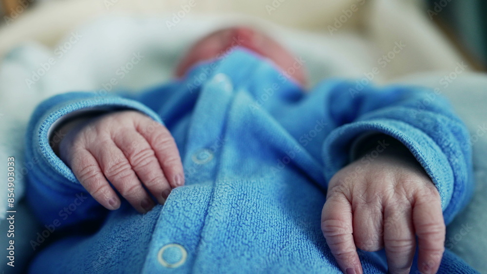 Newborn baby in a blue onesie lying in a crib, focus on tiny hands ...