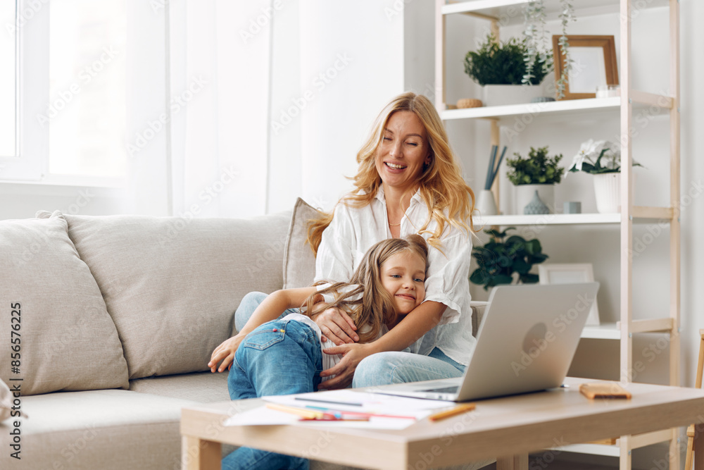 Mother working on laptop with daughter on her lap, cozy living room setting with couch and home ...