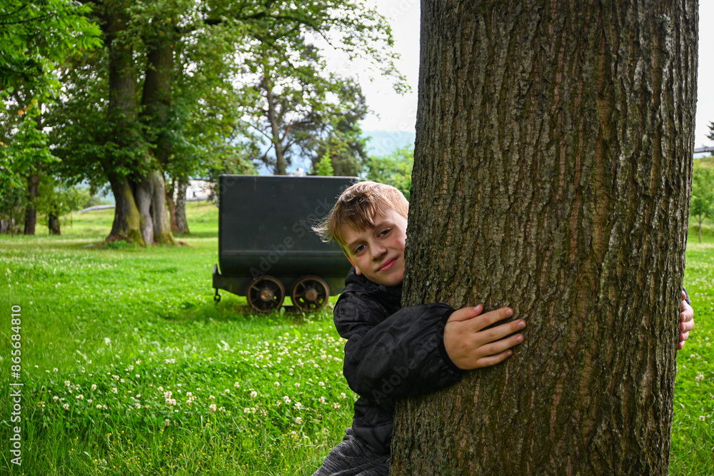 A young boy hugs a big tree in a grassy park and expresses his ...