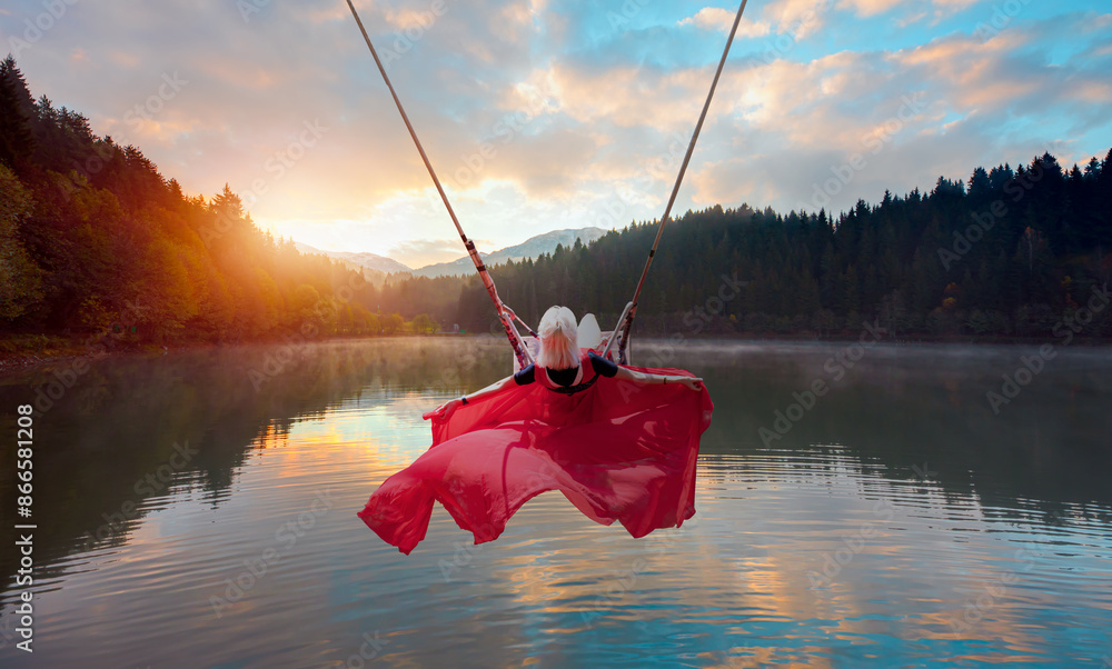 Beautiful blonde woman in a long red dress is swinging on a swing ...