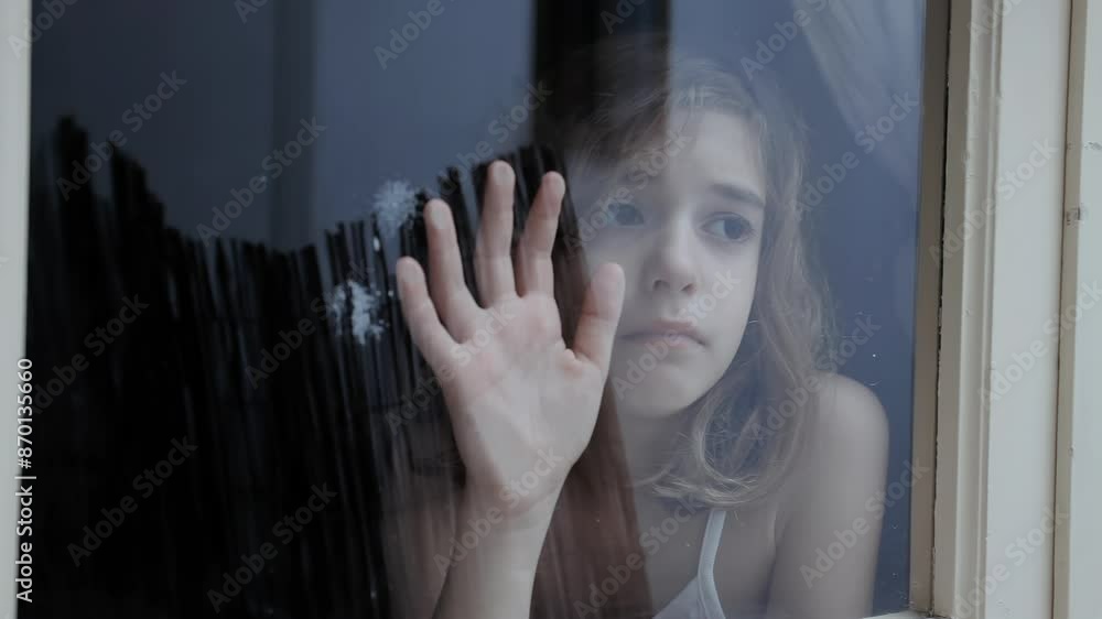 A sad little girl is at home, looking out the window. Her hand touches ...