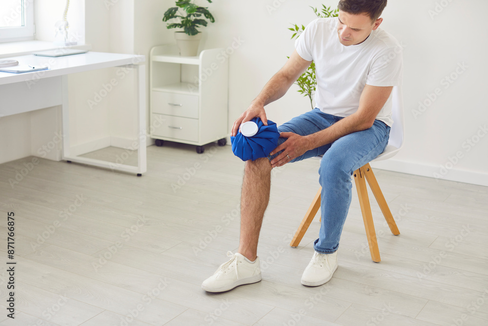 Portrait of a young man applying cold compress to leg sitting on chair ...