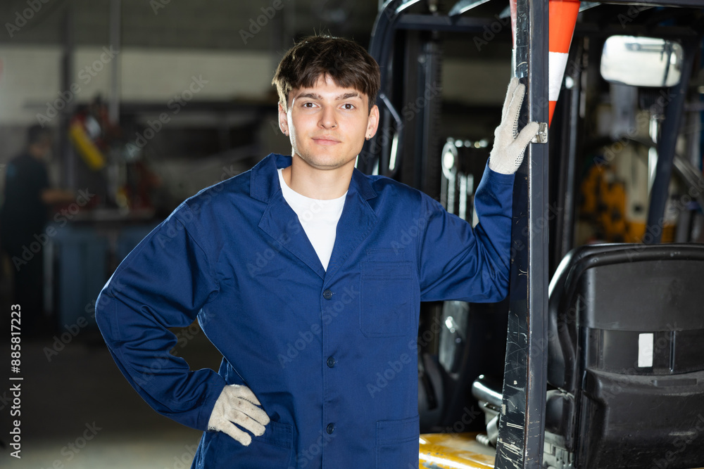 Smiling young male forklift operator in blue work coat and gloves ...