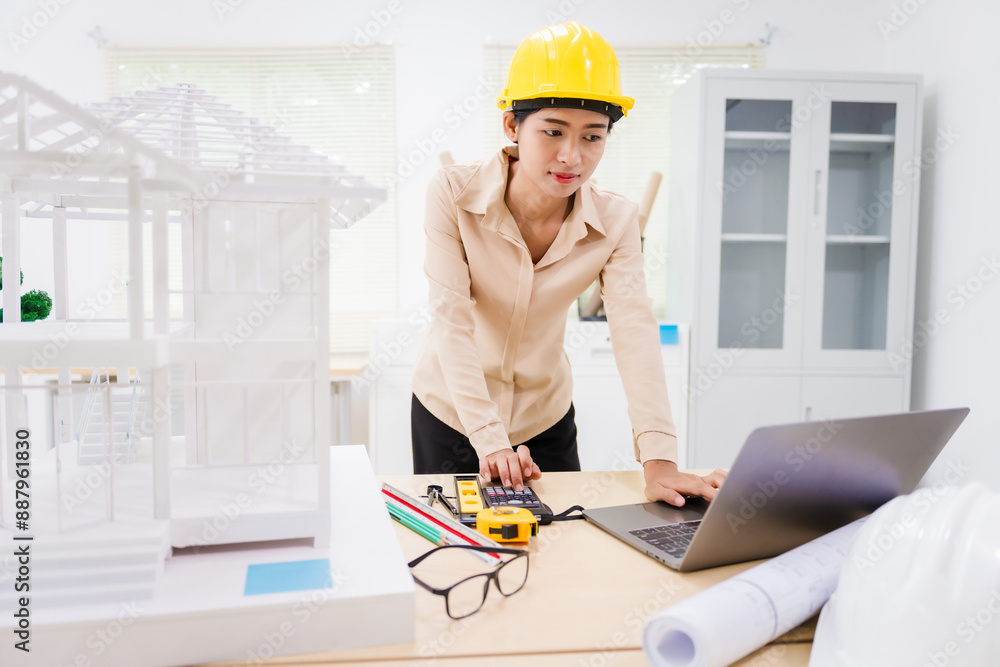 An Asian architect woman sits at her desk, wearing a yellow safety hat ...