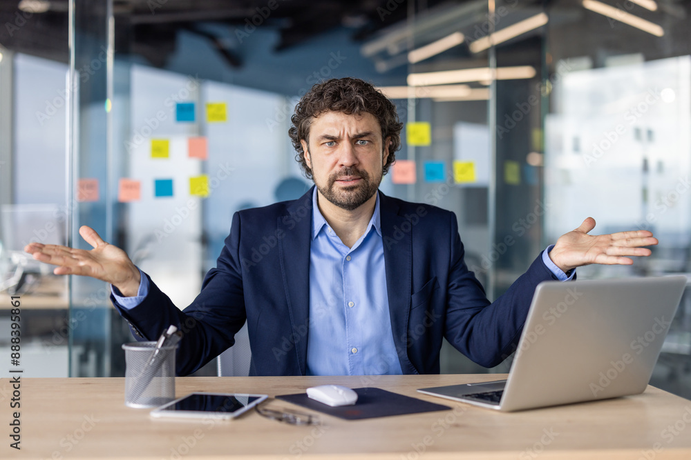 Confused businessman sitting at desk in modern office, shrugging with ...