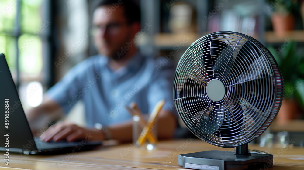 Office worker types on a laptop at a desk, with a cooling fan providing ...
