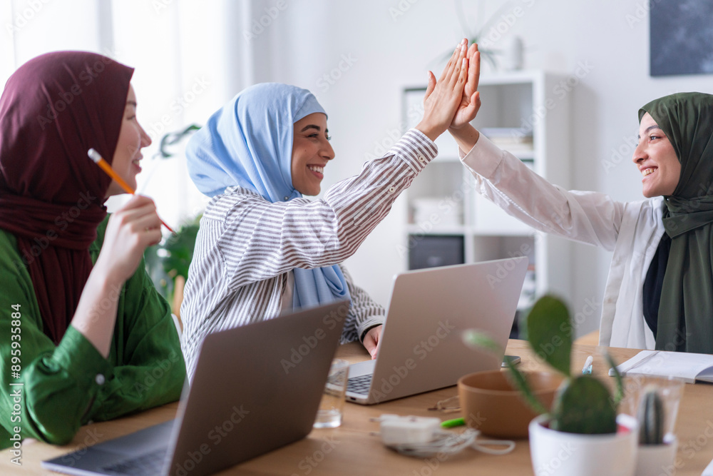 Foto de Three women in hijabs high-five each other in a modern office ...