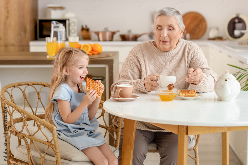 Cute little girl with her granny eating waffle at table in kitchen ...