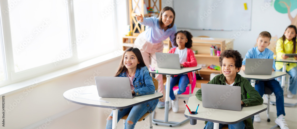 School children using laptops, female teacher pointing at blackboard ...