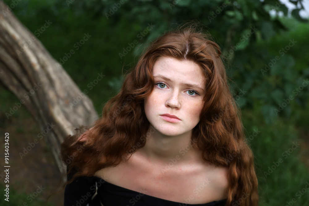 Portrait of upset girl with long red hair and freckles in summer park ...