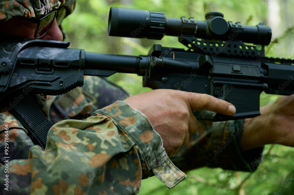 Hunter with rifle wearing camouflage suit sitting in ambush. Camouflaged sniper in the forest in ...