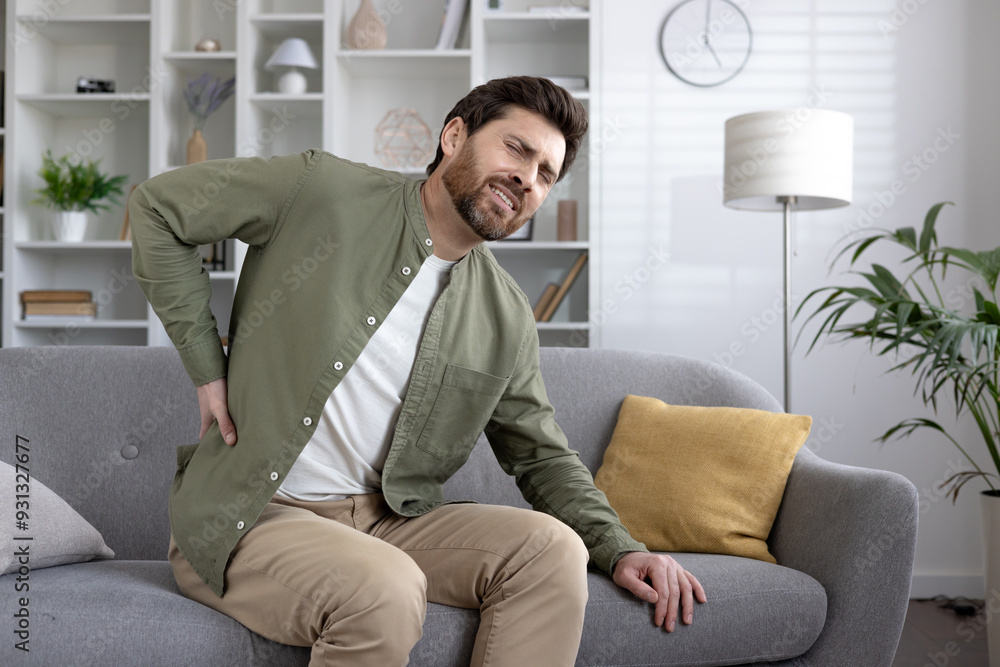 Man seated on couch experiencing back pain, holding lower back. Image conveys discomfort, health ...