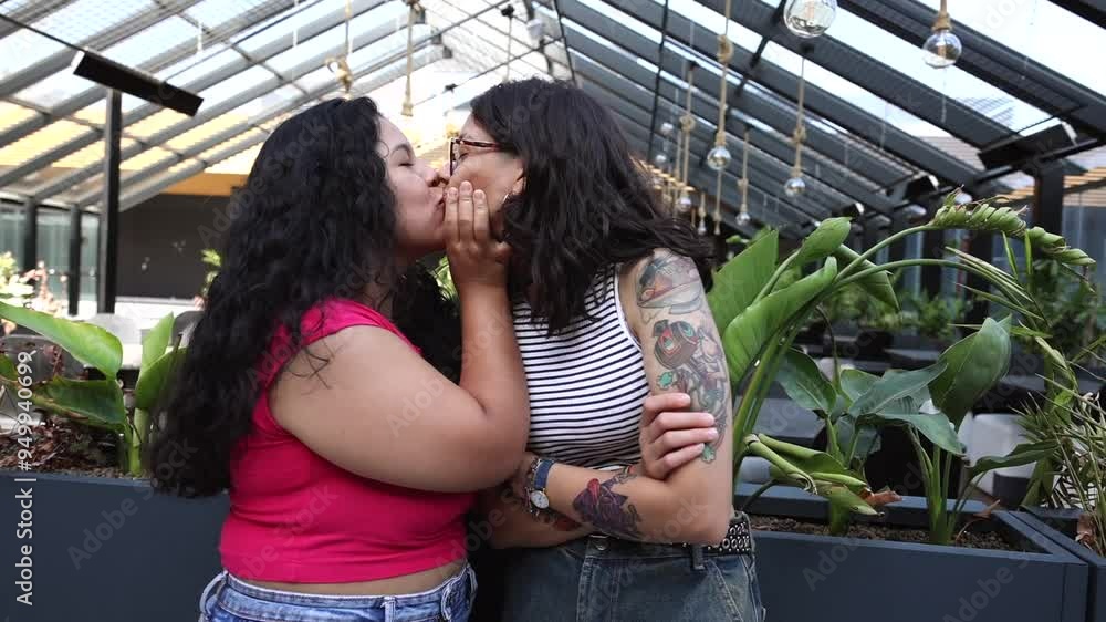 Latin American lesbian couple tenderly kissing in an indoor garden with lush greenery. Latin ...