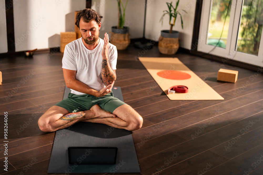 Man practicing yoga at home, sitting on a mat and following an online ...