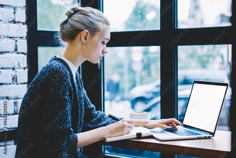Side view of young blonde hair female using laptop with mock up screen ...