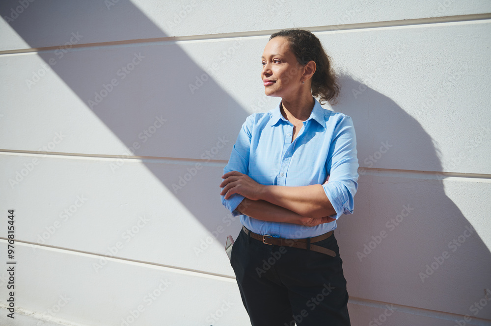 Confident woman in blue shirt posing against a white wall under ...