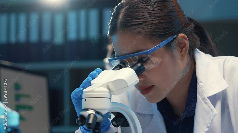 Scientist wearing a lab coat and safety glasses is carefully examining ...