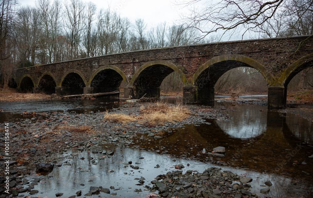 An ancient moss-covered stone arch bridge crosses a quiet river ...