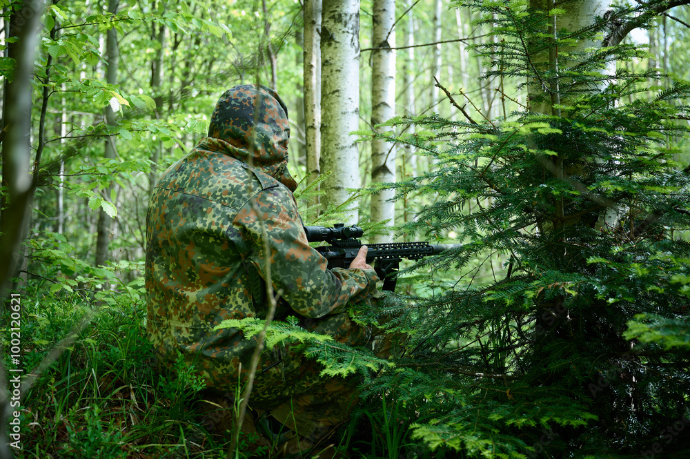 Hunter with rifle wearing camouflage suit sitting in ambush. Camouflaged sniper in the forest in ...